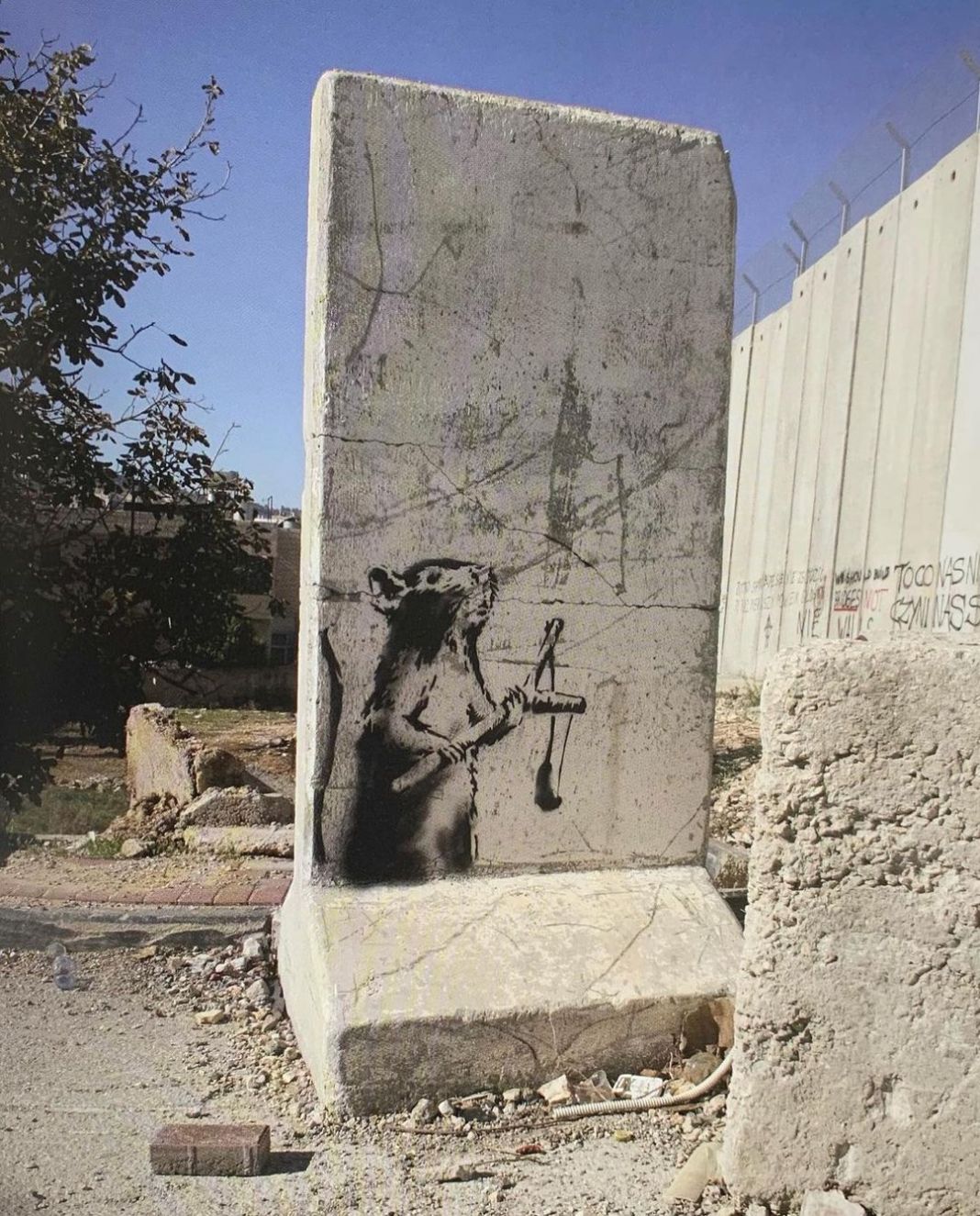 Banksy’s signature rat holds a sling and looks up at the barrier ...